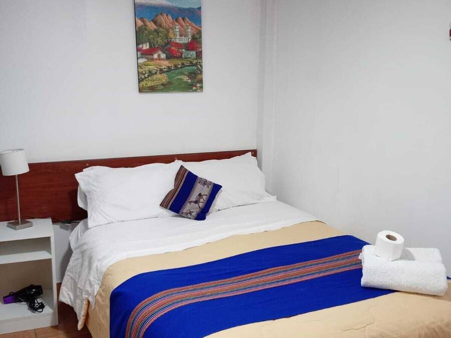 Uma cama bem arrumada com edredom azul e bege, um travesseiro e um quadro de uma paisagem na parede.