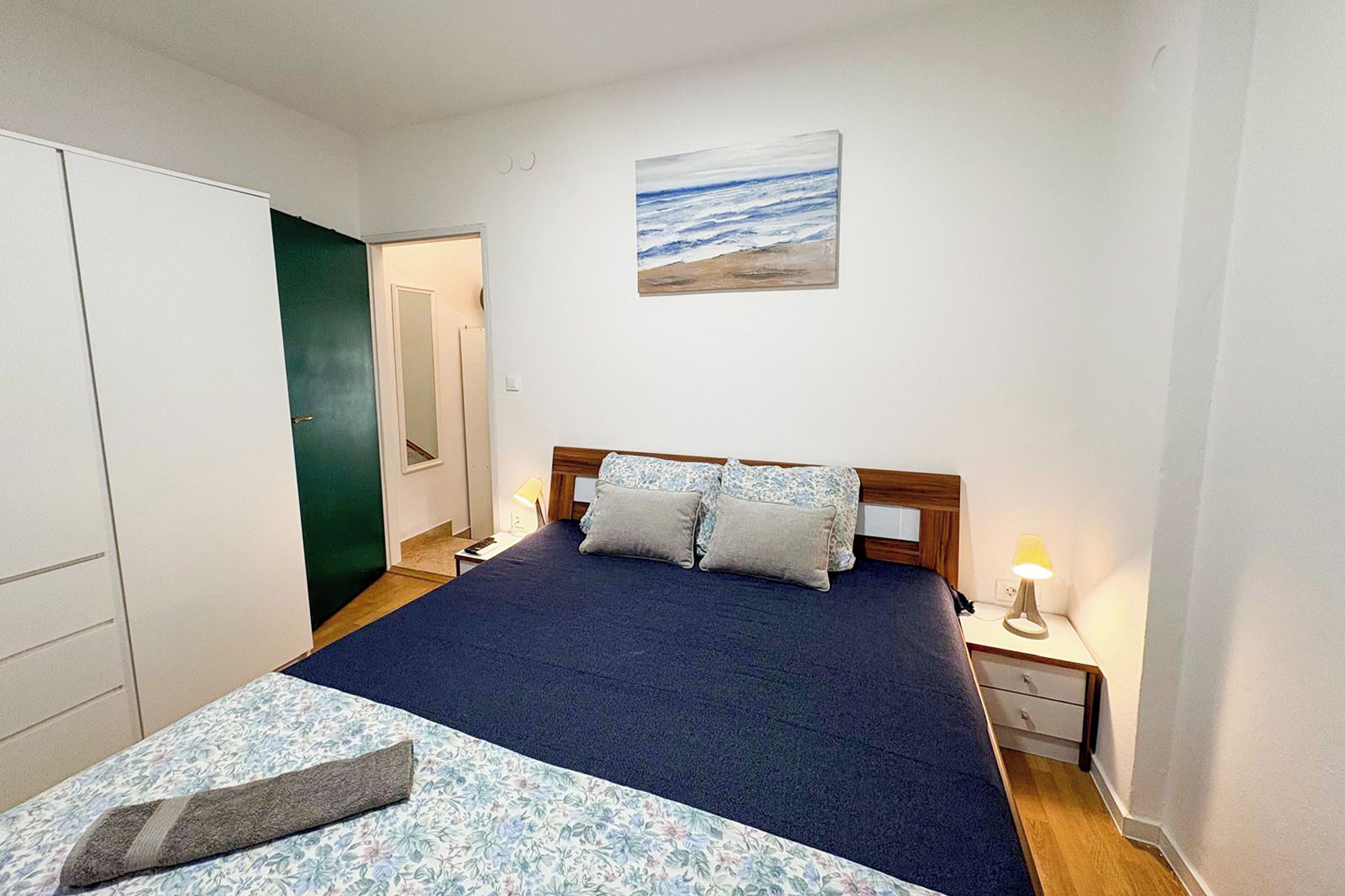 Quarto com cama, duas luminárias de cabeceira, mesa de cabeceira, porta e um quadro na parede.
