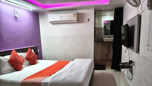 Quarto de hotel com cama, televisão, aparelho de ar condicionado e banheiro visível por uma porta aberta.