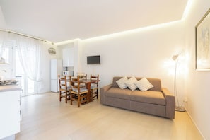 Uma sala de estar moderna com sofá, uma mesa de jantar com cadeiras, uma TV de tela plana na parede e uma cozinha com geladeira e micro-ondas.
