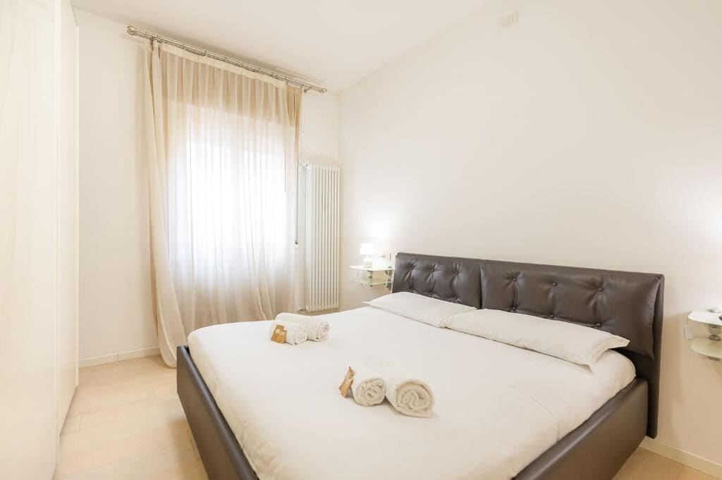Quarto de hotel com uma cama grande, mesa de cabeceira, luminária e janela com cortinas.