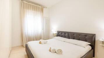 Quarto de hotel com uma cama grande, mesa de cabeceira, luminária e janela com cortinas.