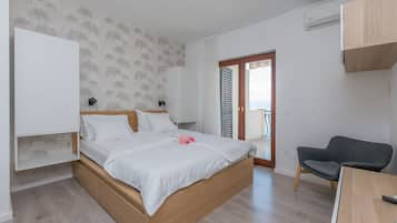 Quarto com cama de madeira, roupa de cama branca, escrivaninha, cadeira e vista através de uma porta.