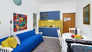 Um espaço de estar compacto com cozinha americana, um sofá azul, uma mesa de jantar com frutas e uma pintura abstrata colorida.