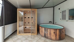 Uma área de spa com uma banheira de madeira, sauna com paredes de vidro e chão ladrilhado.