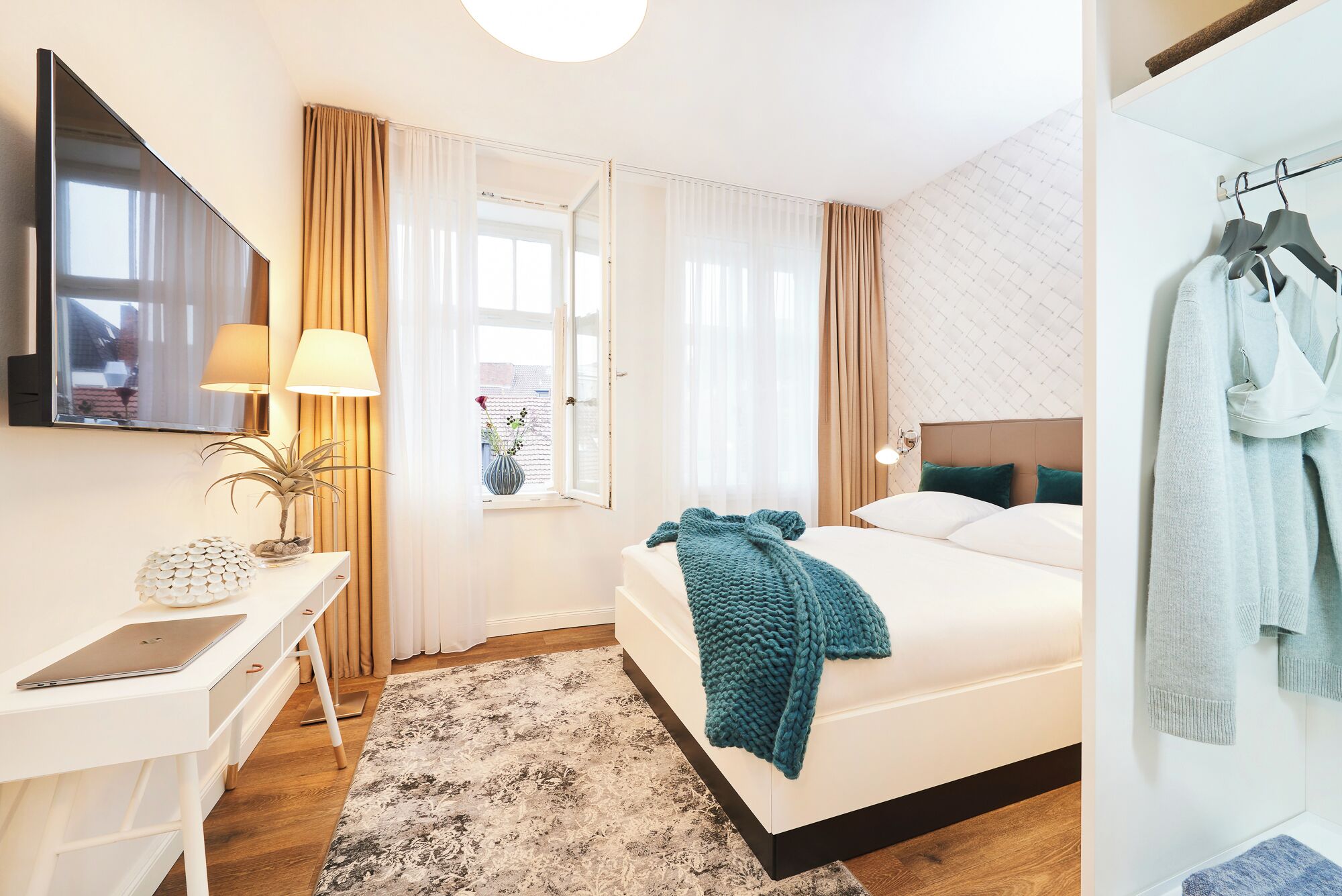 Quarto de hotel com cama, escrivaninha, espelho, janela com cortinas e televisão fixada na parede.