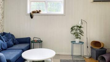 Uma sala de estar com um sofá azul, uma mesa branca, uma planta em vaso, um abajur e um gato no parapeito da janela.