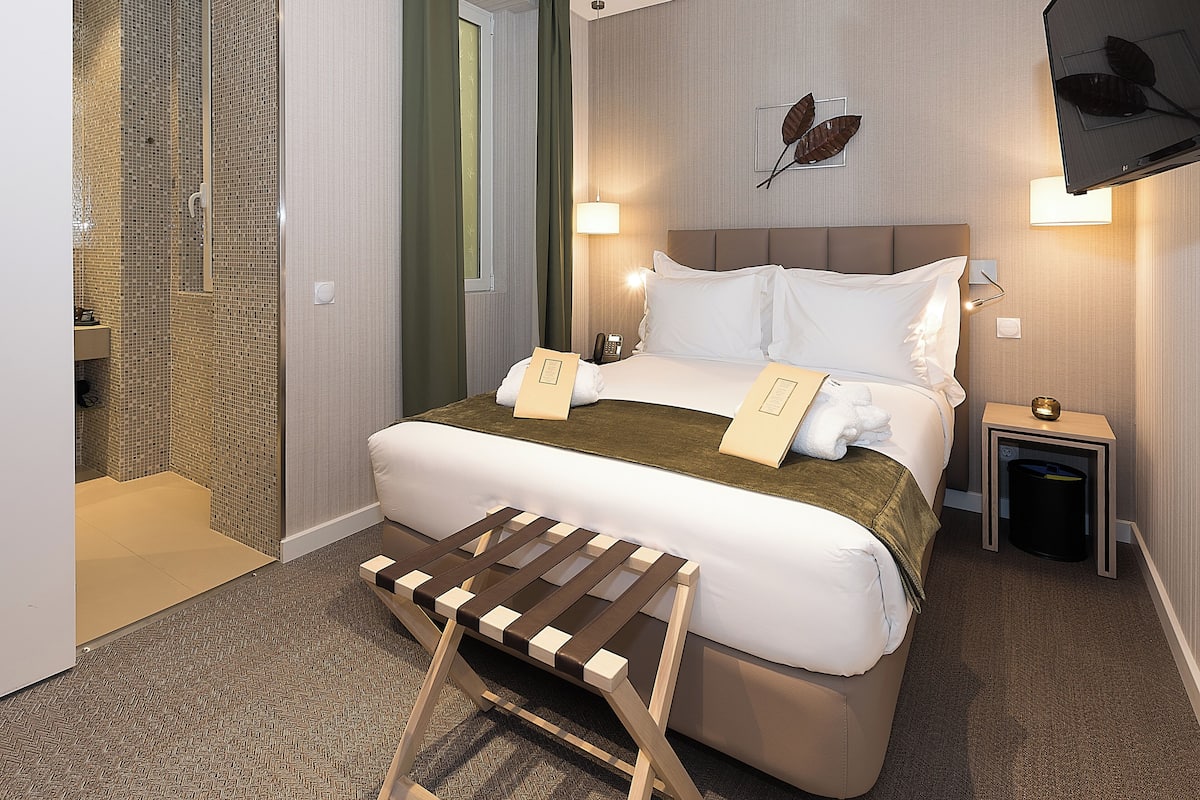 Quarto de hotel com cama, mesa de cabeceira, luminária, televisão e área de chuveiro.
