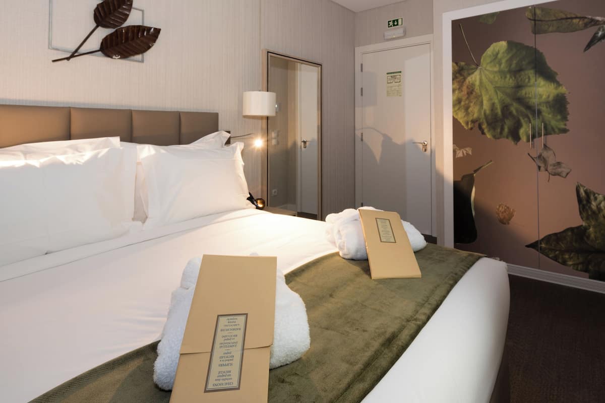 Quarto de hotel com uma cama, mesa de cabeceira, abajur, porta e parede com um mural de folhas.
