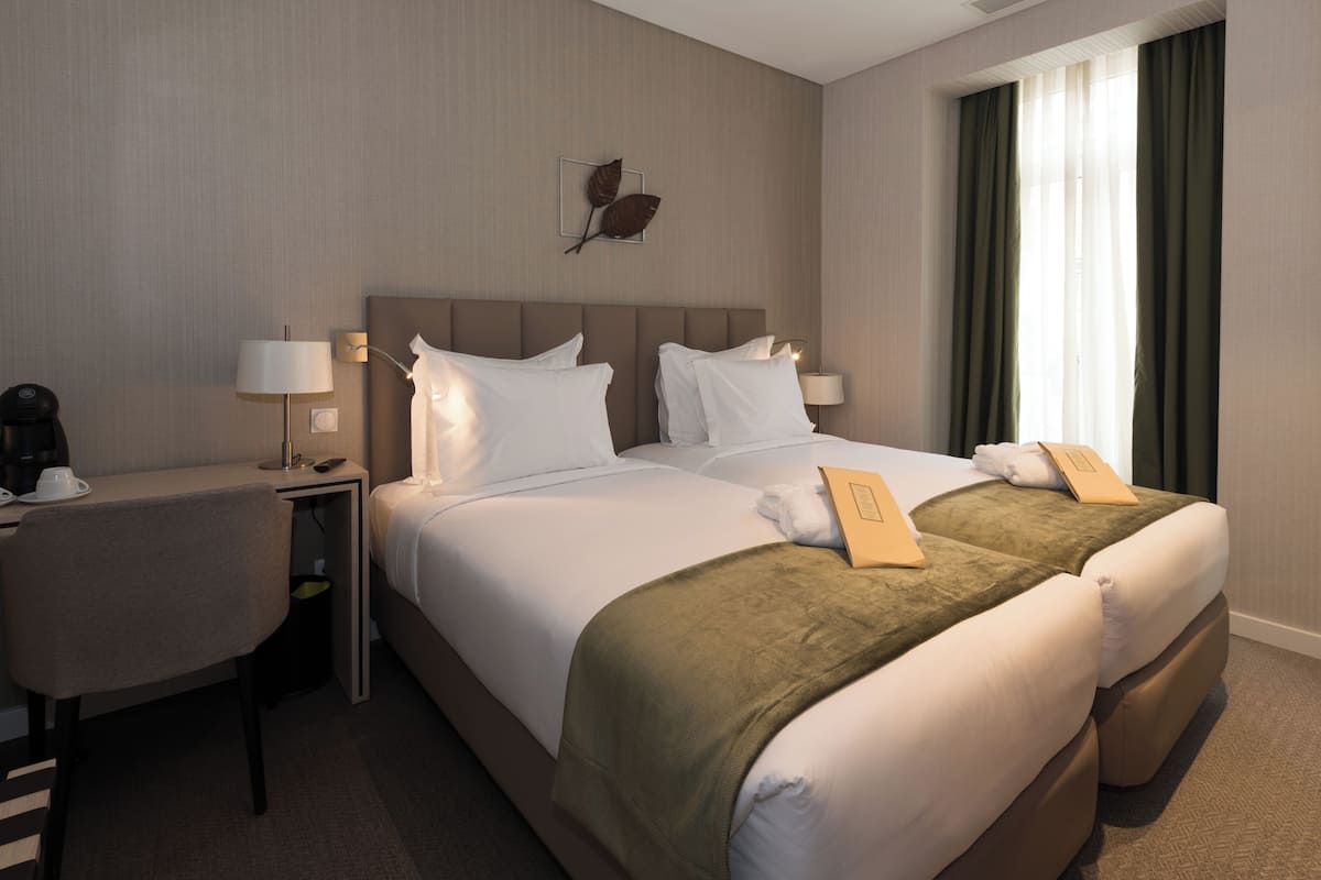 Quarto de hotel com duas camas, uma escrivaninha, uma cadeira, uma cafeteira e uma luminária na parede.