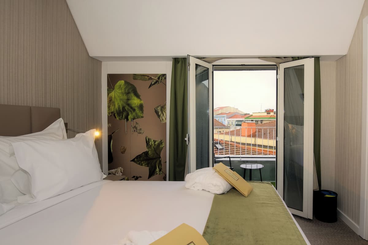 Quarto de hotel com cama, tapete verde, vista de edifícios através de uma porta de vidro e uma pequena mesa com uma cadeira.