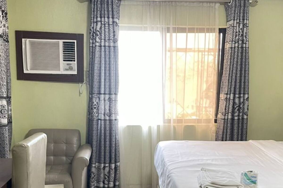 Quarto de hotel com cama, cadeira, escrivaninha, ar condicionado e cortinas.