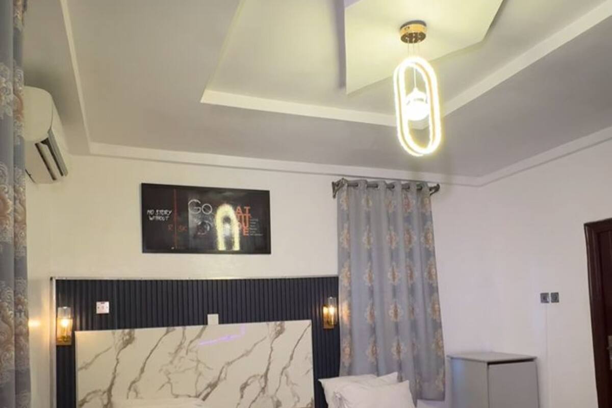 Quarto moderno com cama grande, cabeceira com padrão marmorizado, mesa de cabeceira e luminária no teto.