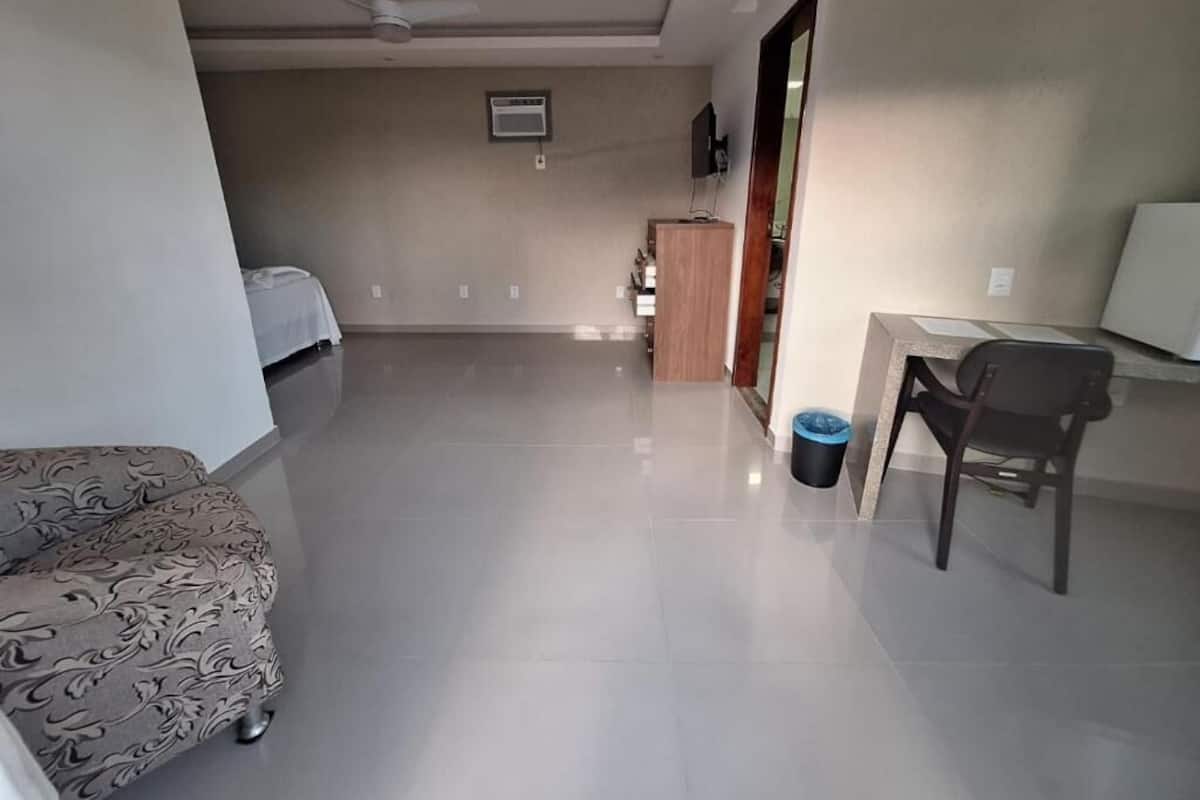 Quarto de hotel com cama, escrivaninha, cadeira, lixeira, ventilador de teto e televisão.