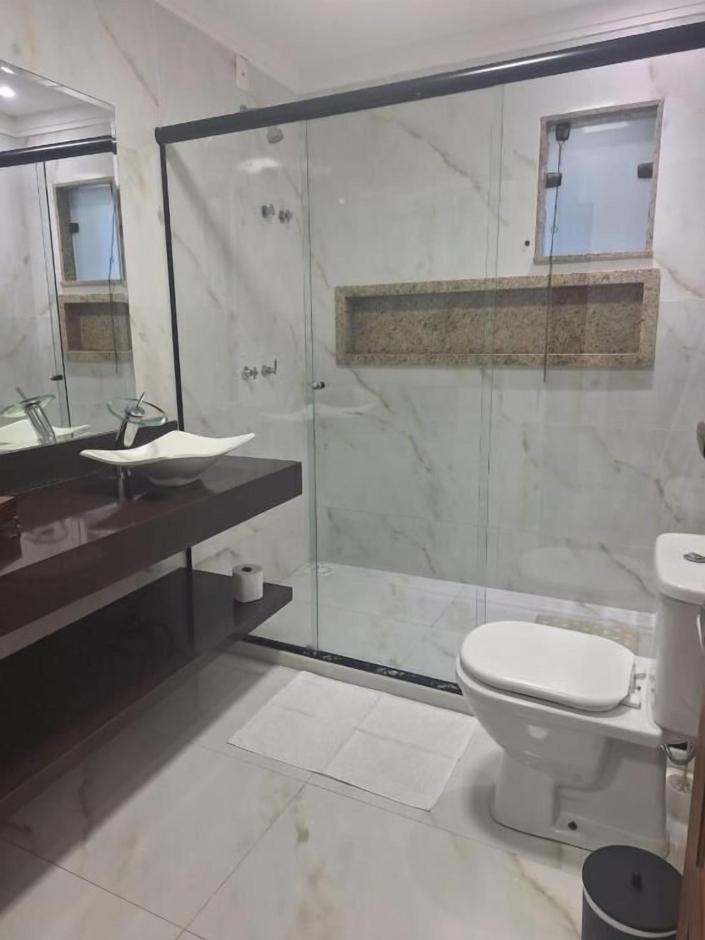 Uma casa de banho moderna com cabine de duche em vidro, soalho em mármore, bancada escura e sanita branca.