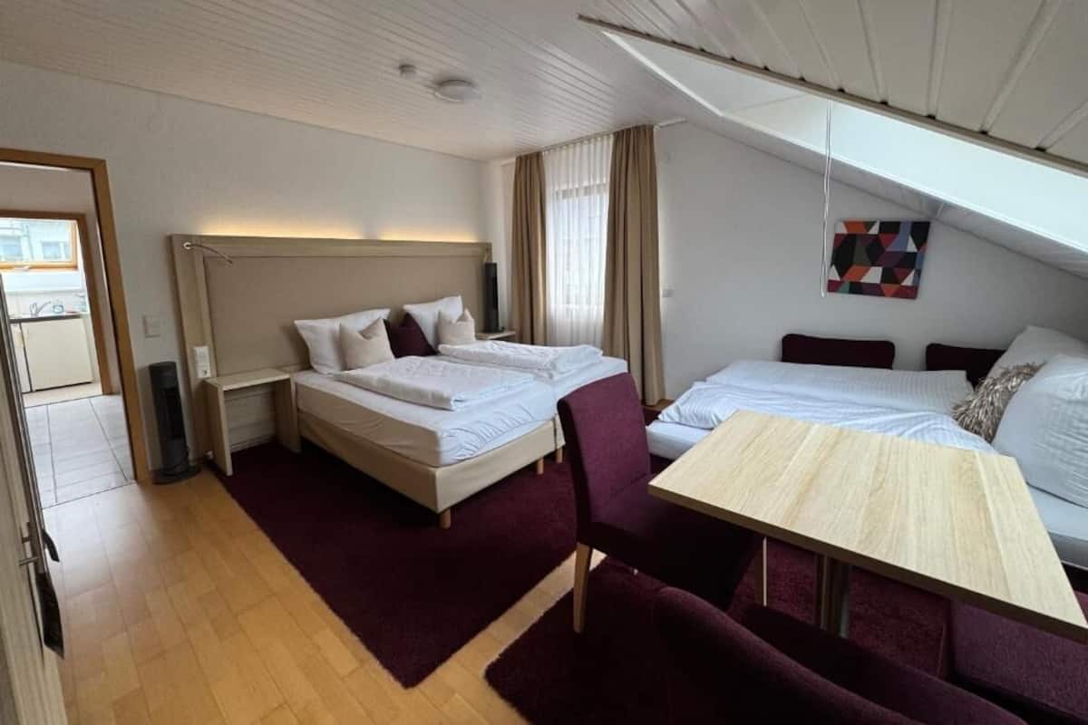 Quarto de hotel com duas camas, uma mesa de madeira, uma cadeira vermelha e vista para o exterior.