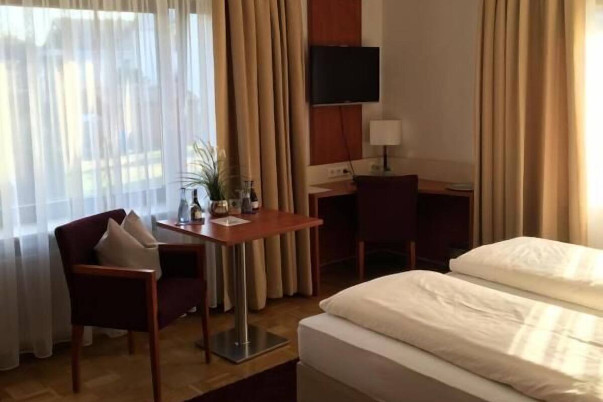 Quarto de hotel com cama, escrivaninha, cadeira, televisão e cortinas.