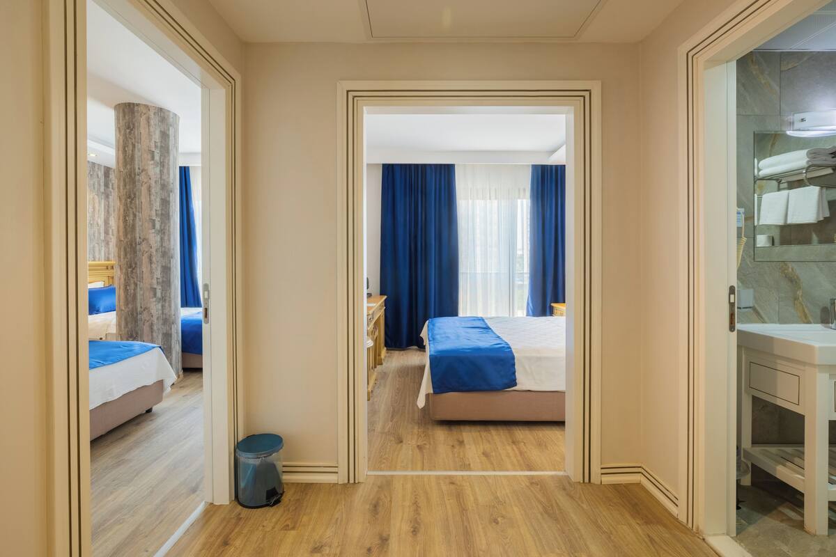 Quarto de hotel com piso de madeira, cama com colcha azul, escrivaninha com cadeira, espelho e banheiro com pia e porta-toalhas.
