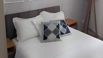 Cama bem arrumada com roupa de cama branca, um travesseiro decorativo e uma parede com arte abstrata. Há duas mesas de cabeceira e um abajur.