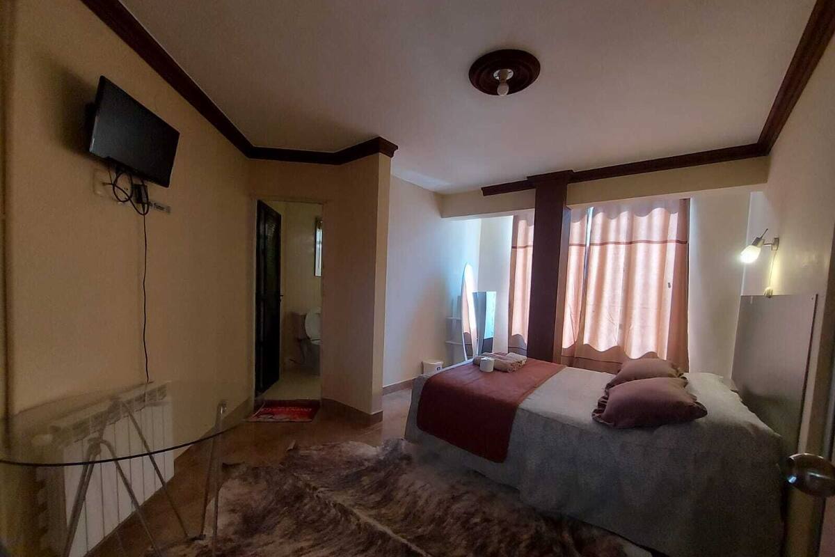 Quarto com cama, televisão na parede, espelho e janela com cortinas.