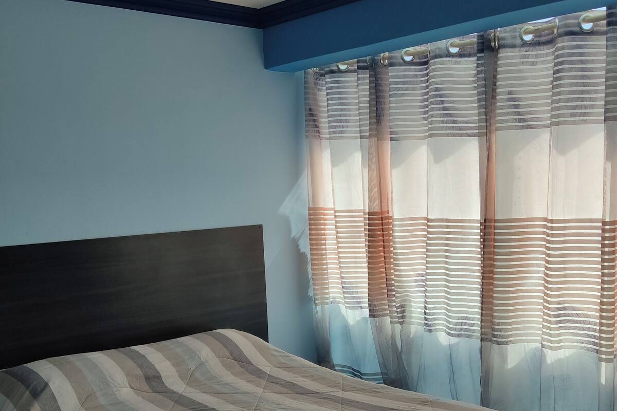 Quarto com cama, cabeceira e janela com cortinas.