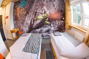 Quarto com duas camas, painel de parede com temática de escalada, uma mesinha e uma janela com cortinas.