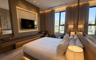 Quarto de hotel moderno com uma cama grande, televisão de tela plana na parede, escrivaninha e vista para a cidade através de amplas janelas.