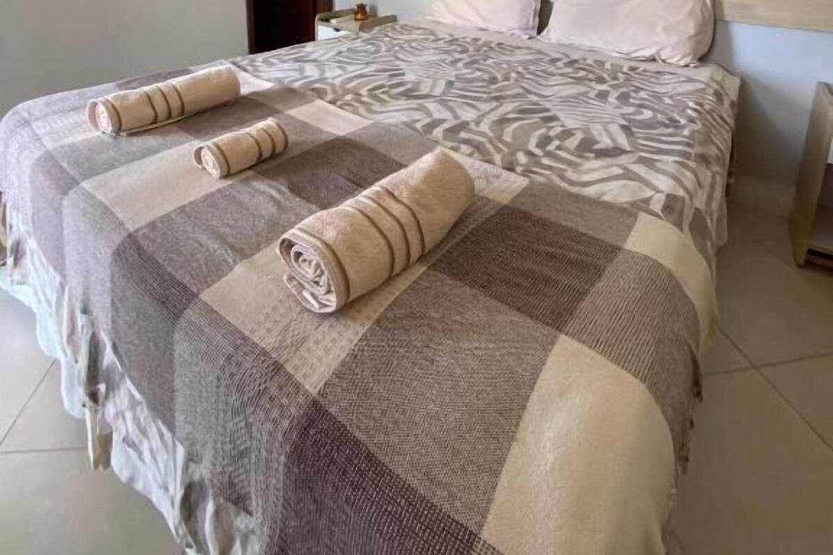 Cama bem arrumada com toalhas dobradas, cabeceira, quadro na parede e porta à esquerda.