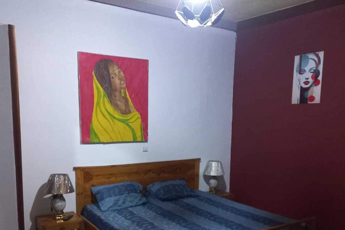 Quarto com cama de madeira, mesa de cabeceira com abajur, uma cadeira e duas pinturas na parede.
