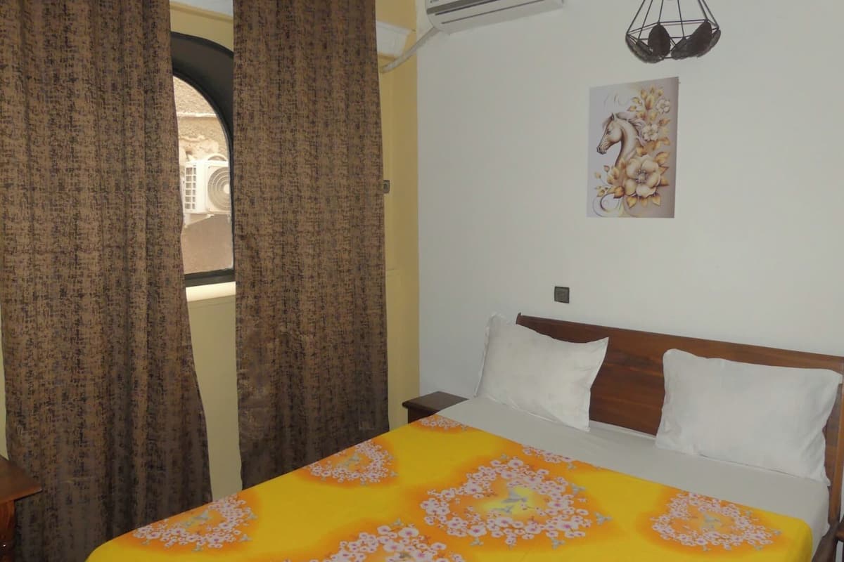 Quarto com cama, cortinas, ar condicionado, luminária fixada na parede e um quadro com flores.