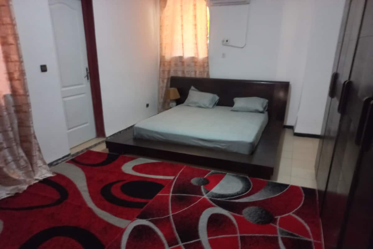 Quarto com uma cama, um tapete estampado vermelho, uma porta branca e uma parede branca.