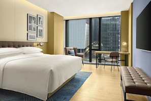 Quarto de hotel com uma cama grande, uma escrivaninha, uma cadeira e vista para a cidade através de grandes janelas.