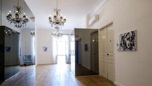 Um corredor amplo com piso de madeira, um candelabro, um espelho e uma porta que dá para um quarto.