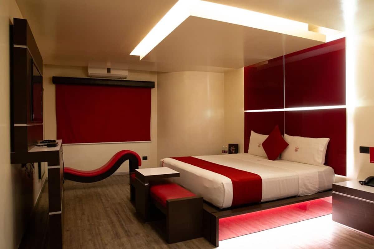 Quarto de hotel moderno com cama, mesa de cabeceira, escrivaninha, cadeira e uma parede de destaque vermelha.