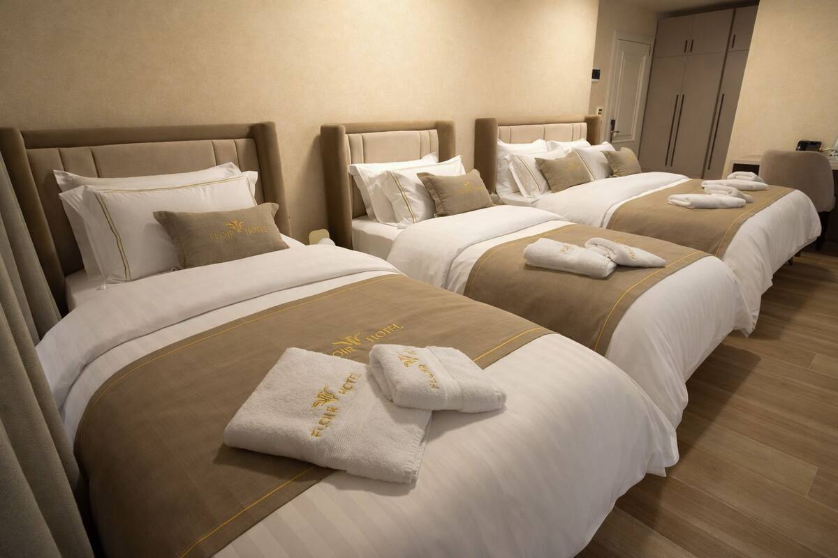 Quarto de hotel com três camas, cada uma com cabeceira, roupa de cama branca e detalhes em castanho. Há toalhas nas camas e um guarda-roupa ao fundo.