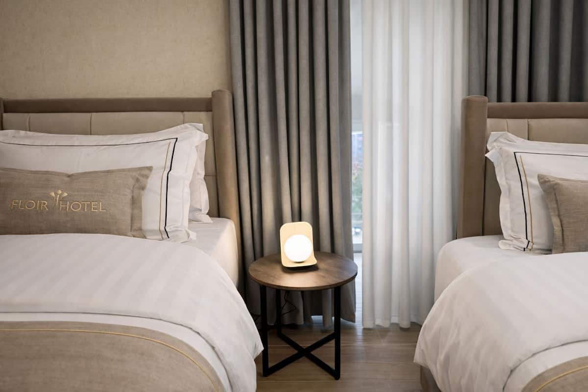 Quarto de hotel com duas camas, uma mesa pequena com abajur e vista através de cortinas translúcidas.