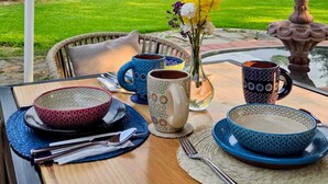 Mesa posta com pratos, tigelas e canecas, um vaso com flores e uma fonte em um jardim com bancos e montanhas ao fundo.