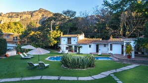 Uma área de piscina com espreguiçadeiras e guarda-sóis, rodeada por um gramado bem cuidado e um edifício branco com telhado em azulejo.