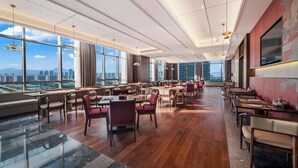 Um restaurante amplo com pé-direito alto, grandes janelas e vista para a cidade.