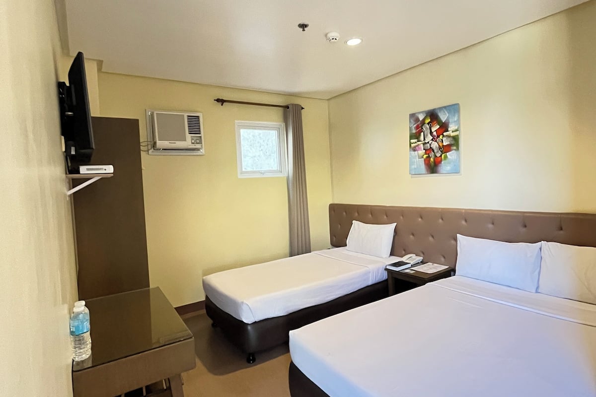 Quarto de hotel com duas camas, uma escrivaninha, televisão, aparelho de ar condicionado, janela e um quadro na parede.