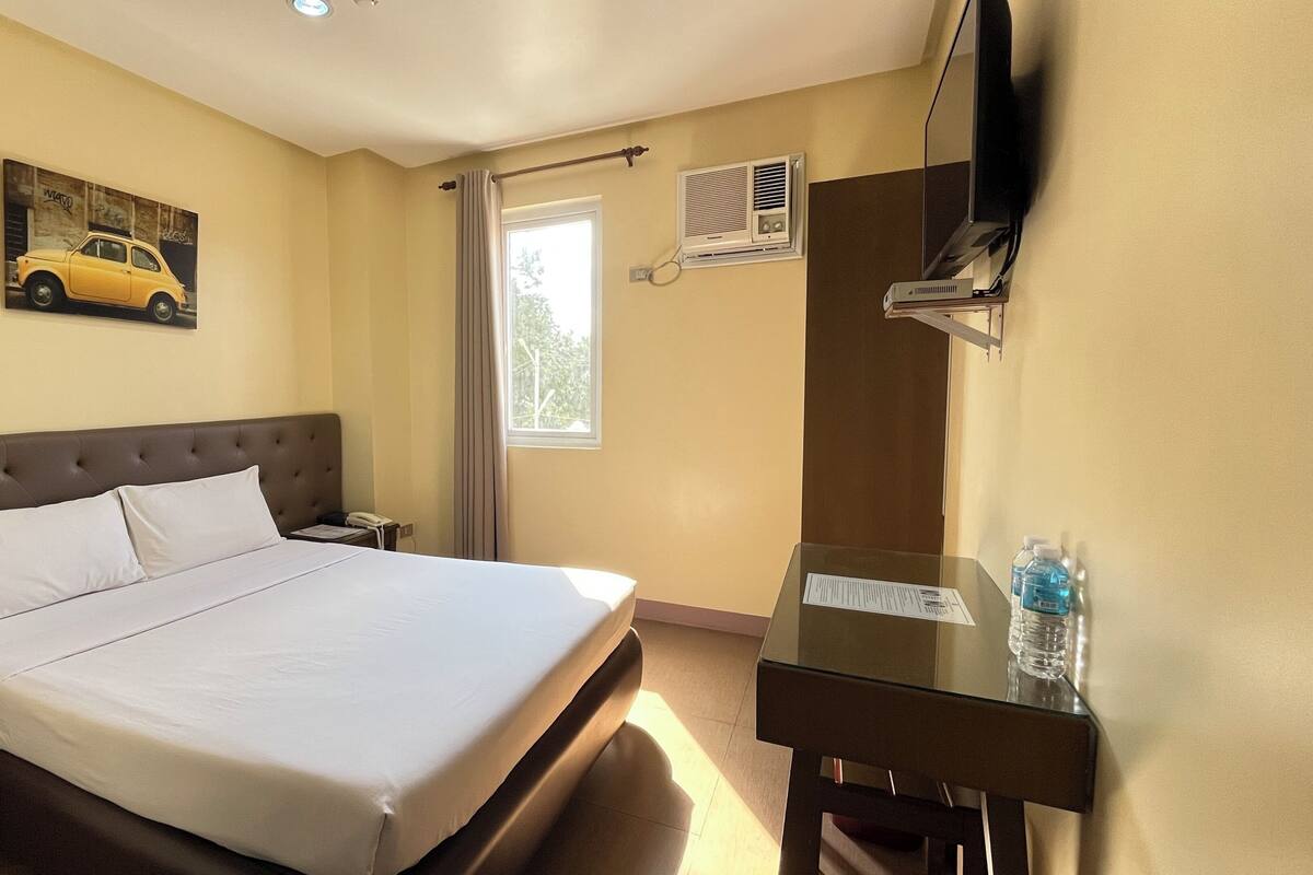 Quarto de hotel com cama, mesa de cabeceira, escrivaninha com espelho, janela com cortinas e ar condicionado.