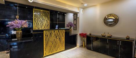 Uma área de recepção com balcão de mármore preto, um painel decorativo amarelo e um espelho na parede.