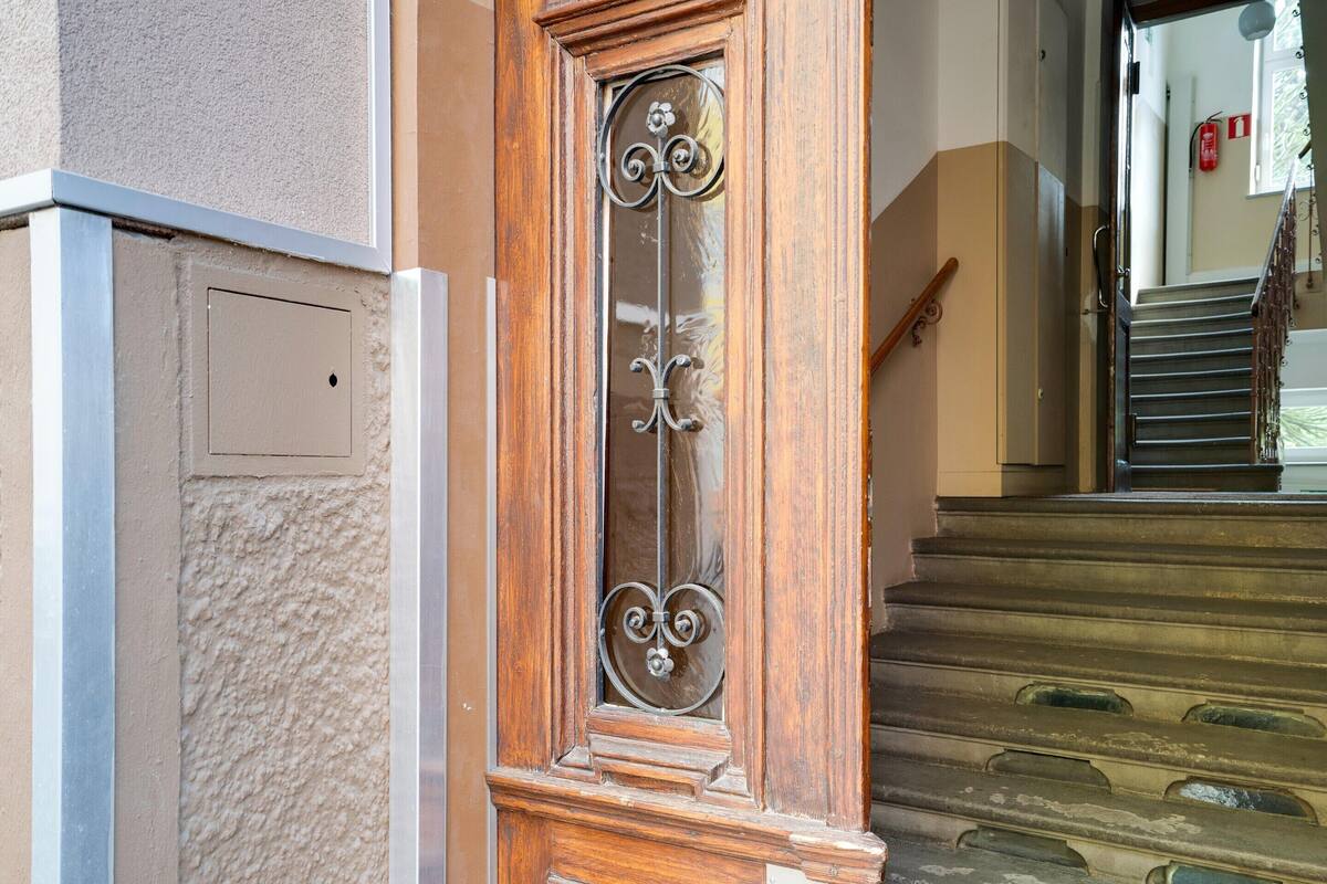 Uma porta de madeira com painel de vidro decorativo, parede texturizada e escada com corrimão de metal.