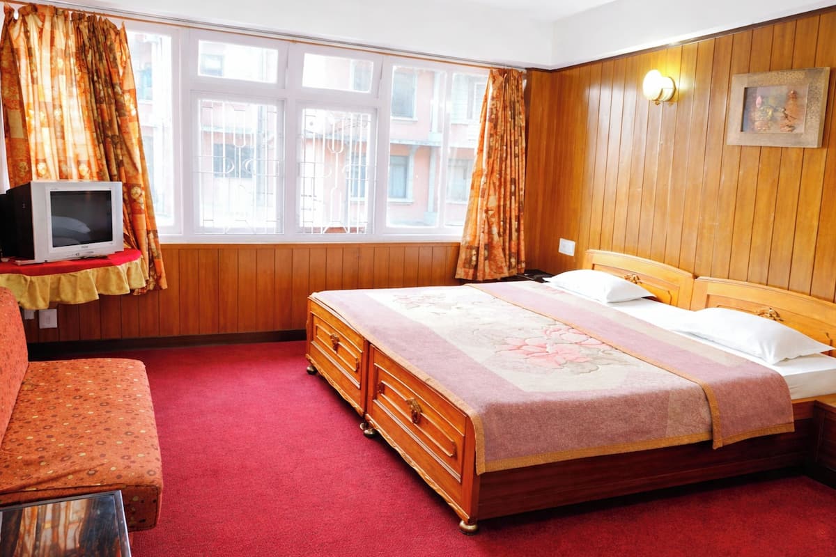 Quarto com cabeceira de madeira, cama com colcha florida, televisão sobre uma mesinha e janela com cortinas.