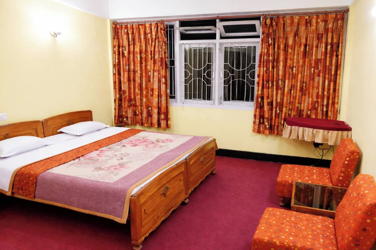 Um quarto com cama, cómoda de madeira, cadeira, janela com cortinas e uma pequena mesa.