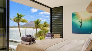 Quarto com uma janela grande, uma cama e vista para uma praia e palmeiras.