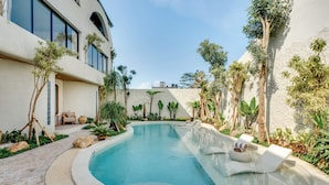 Piscina com espreguiçadeiras, rodeada por plantas tropicais e um edifício moderno com grandes janelas.