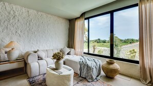 Uma sala de estar moderna com um sofá, uma mesinha lateral, um abajur, um vaso com flores, um tapete e uma janela grande com vista.