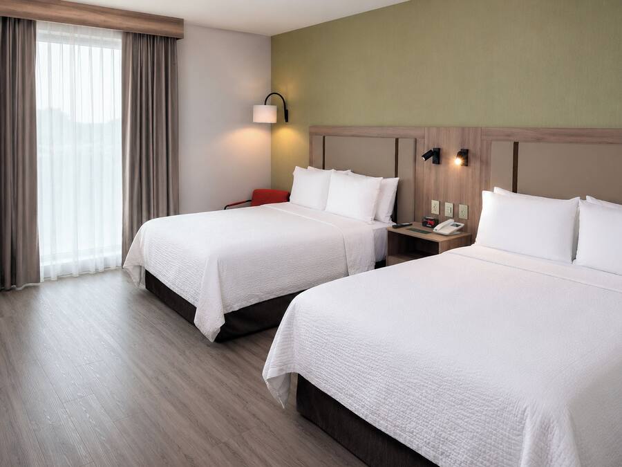 Quarto de hotel com duas camas, mesa de cabeceira com abajur e vista para o exterior pela janela.