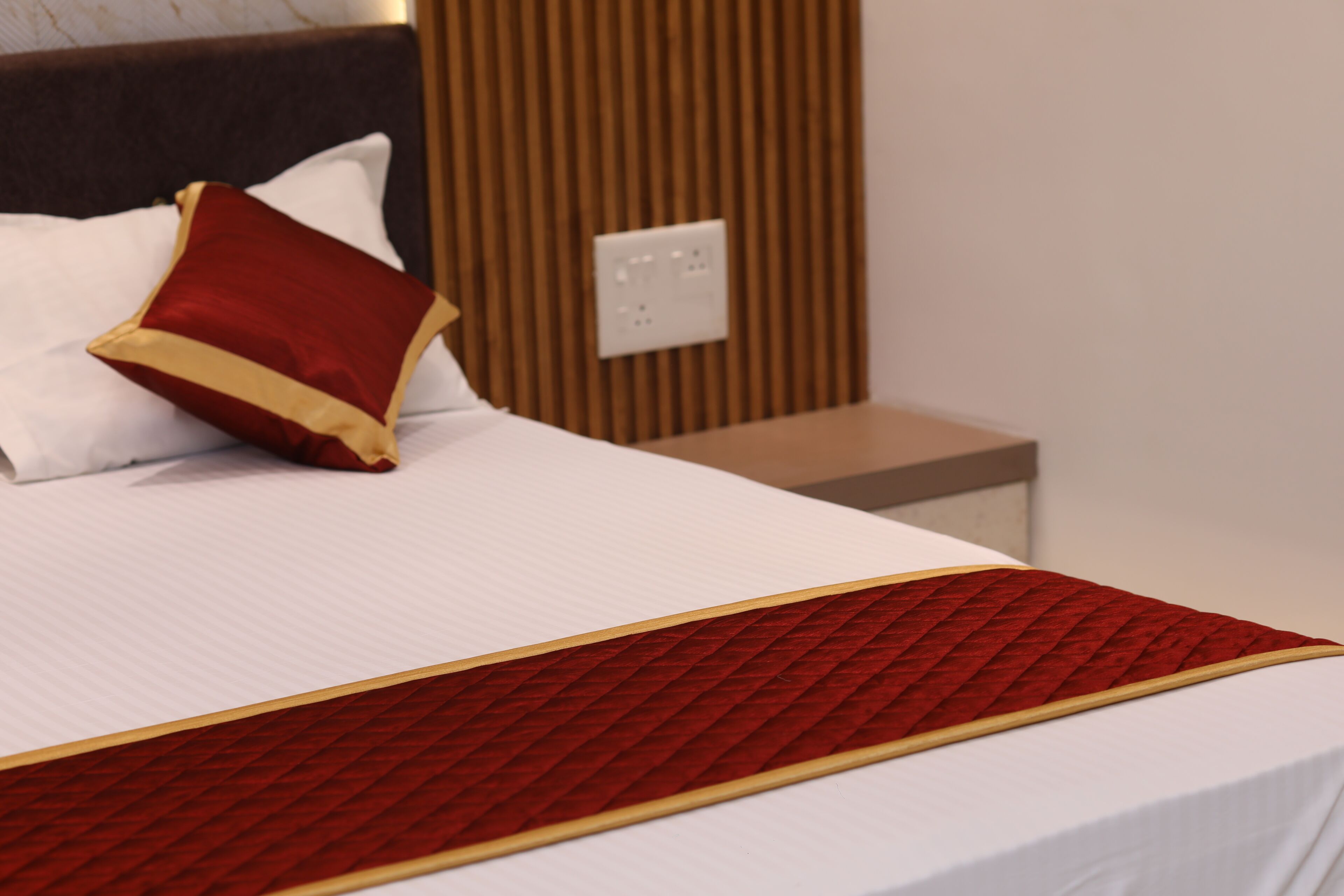 Uma cama bem arrumada com travesseiro de destaque em vermelho e dourado, colcha branca e cabeceira de madeira.